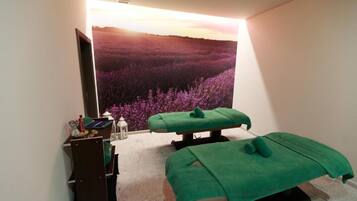 Um quarto com duas mesas de massagem verdes, um pequeno armário e uma grande tapeçaria na parede retratando um campo roxo.
