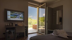 Quarto de hotel com cama, escrivaninha, televisão e varanda com vista para o campo.