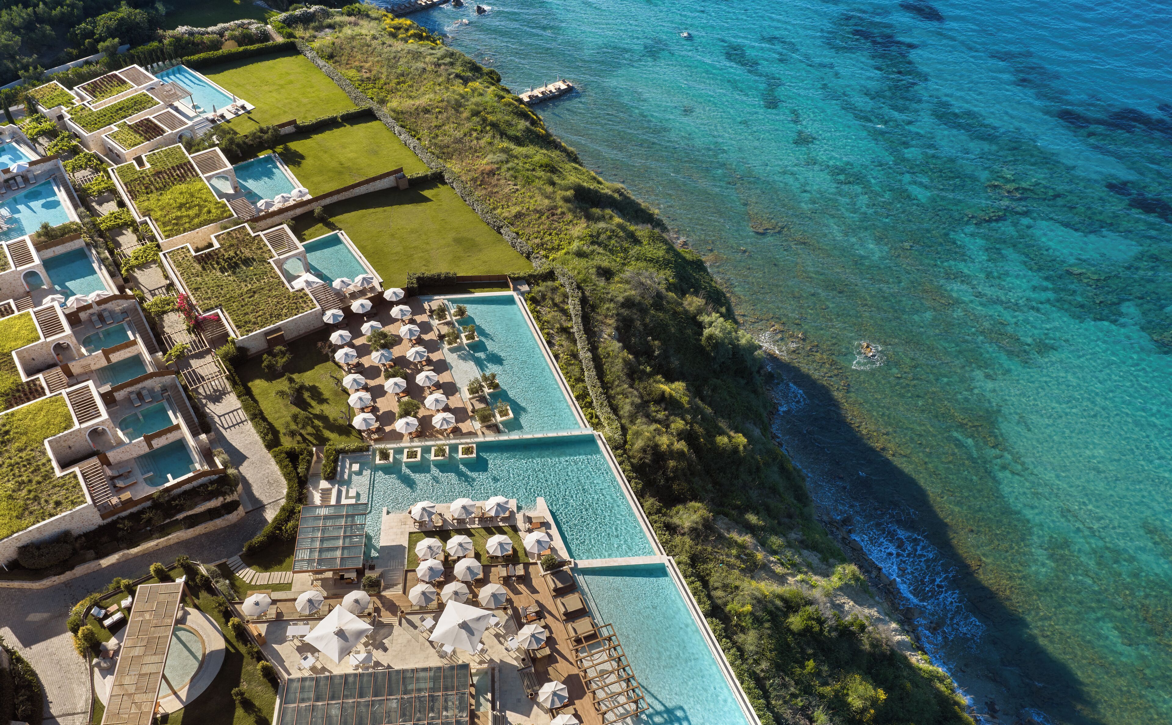 Vista aérea de um resort com várias piscinas, espreguiçadeiras e uma costa rochosa.