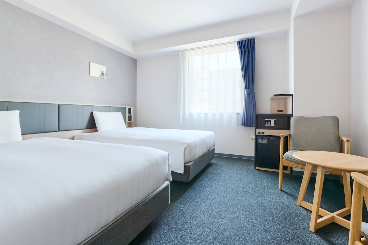 Quarto de hotel com duas camas, uma escrivaninha, uma cadeira, um pequeno armário e uma janela com cortinas.