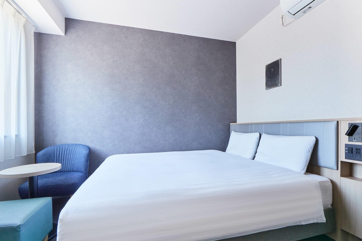 Quarto de hotel com uma cama grande, uma mesa redonda pequena, uma cadeira azul e uma parede cinza decorativa.