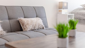 Uma sala de estar moderna com um sofá cinzento, uma almofada fofa, uma mesa de centro de madeira e duas plantas em vasos.