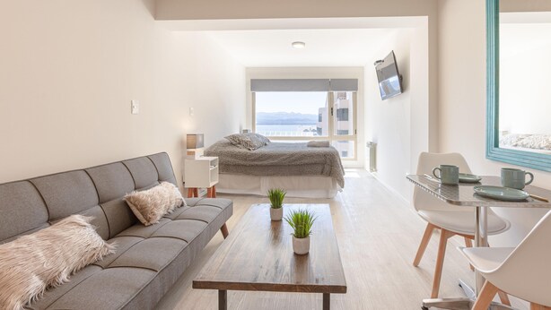 Uma sala de estar moderna e bem iluminada, com um sofá cinzento, uma mesa de centro de madeira e uma área de jantar com uma pequena mesa e cadeiras.