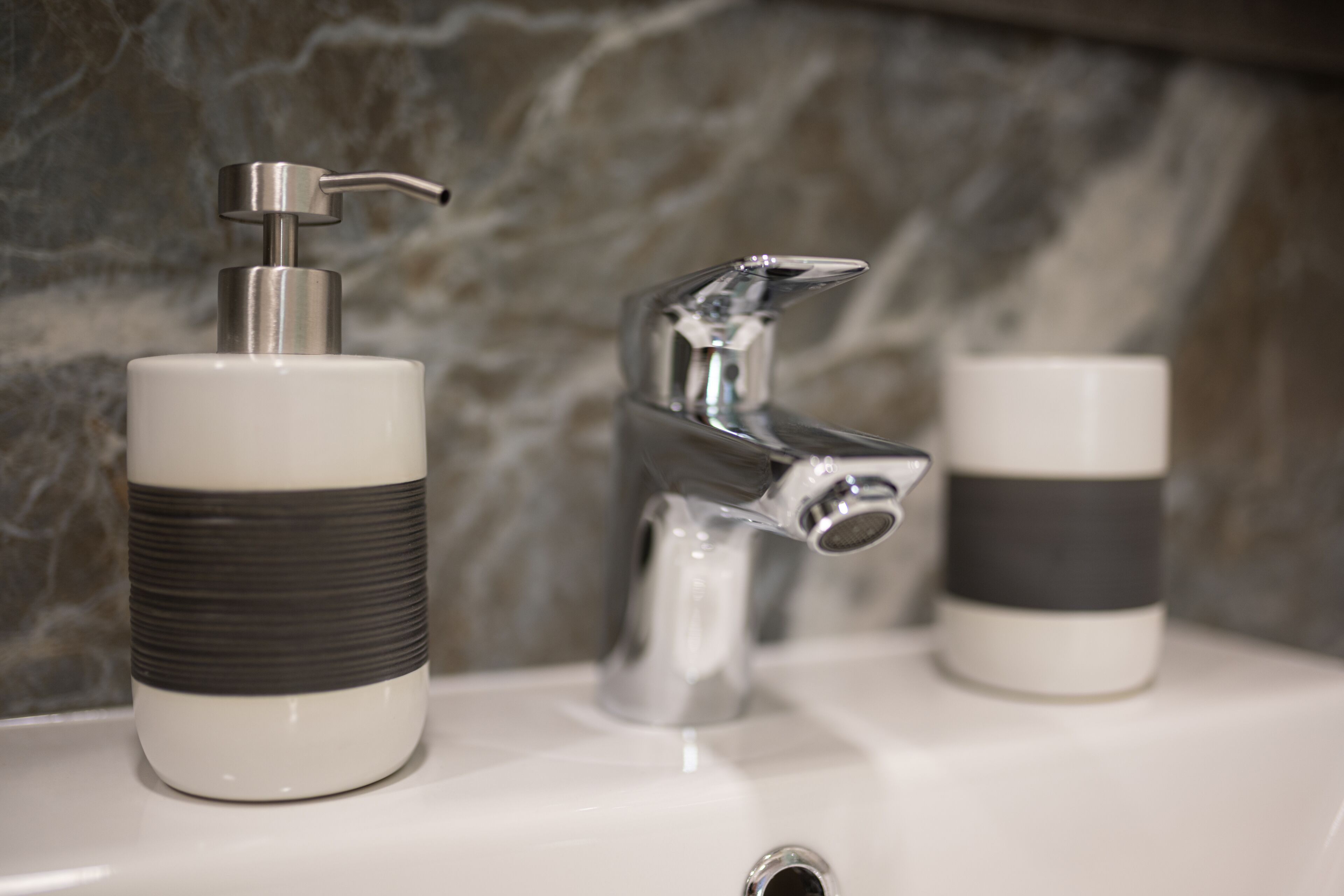 Uma pia de banheiro com uma torneira cromada, um dispensador de sabonete branco com faixas pretas e uma caneca branca com uma faixa preta.