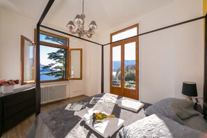 Quarto com uma cama grande, cômoda, radiador, lustre e vista para o mar.