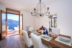 Uma sala de jantar com uma mesa de madeira posta para quatro pessoas, cadeiras brancas, um candelabro e vista para o mar através de uma porta de vidro.