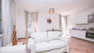 Uma sala de estar com um sofá branco, uma mesa de jantar com cadeiras, uma área de cozinha com armários e eletrodomésticos, e uma janela com cortinas.