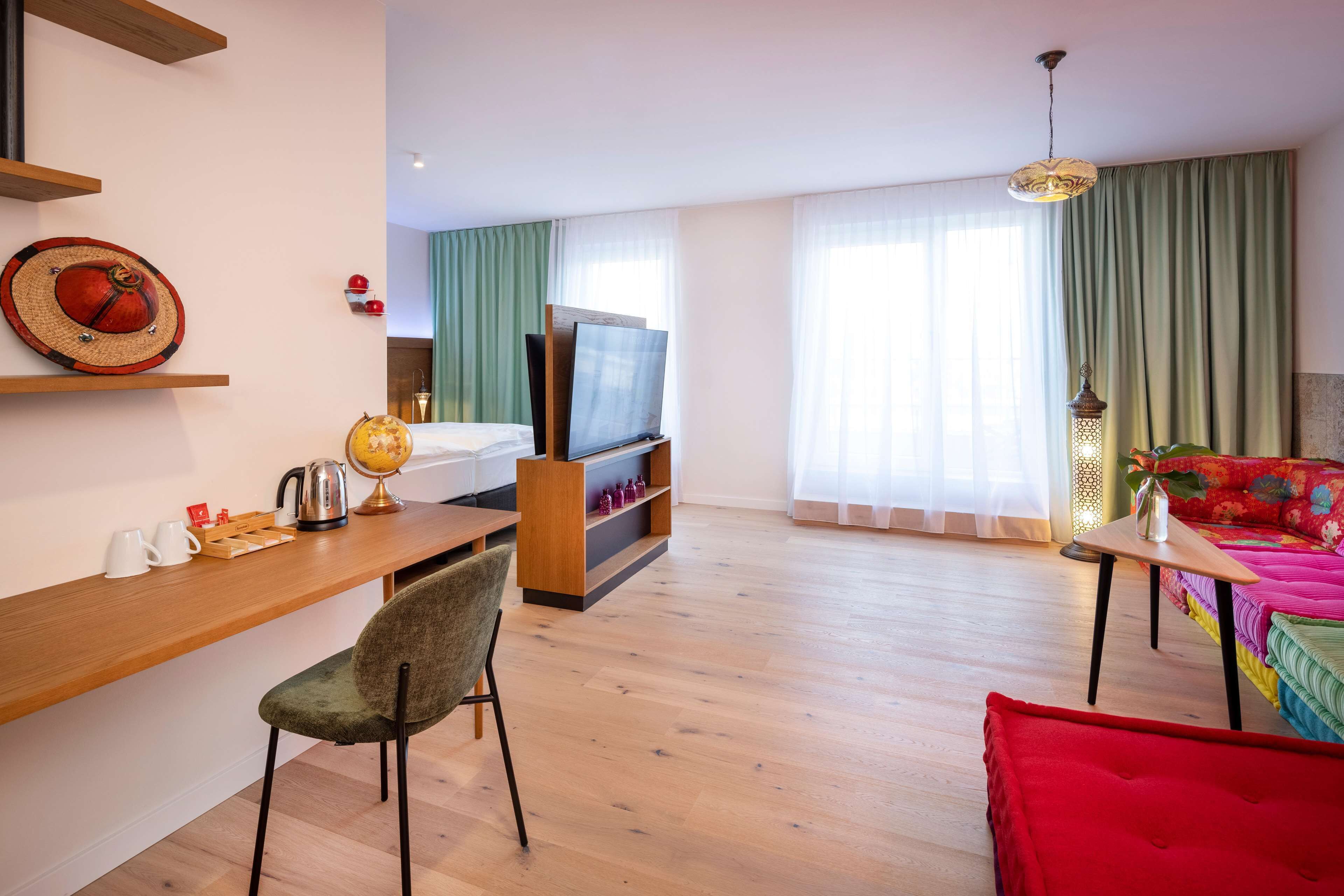 Quarto de hotel moderno com escrivaninha de madeira, uma poltrona confortável, cama com cortinas verdes, televisão e sofá colorido.