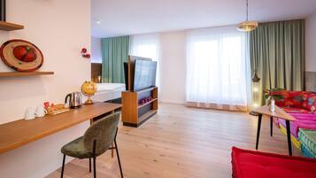 Quarto de hotel moderno com escrivaninha de madeira, uma poltrona confortável, cama com cortinas verdes, televisão e sofá colorido.