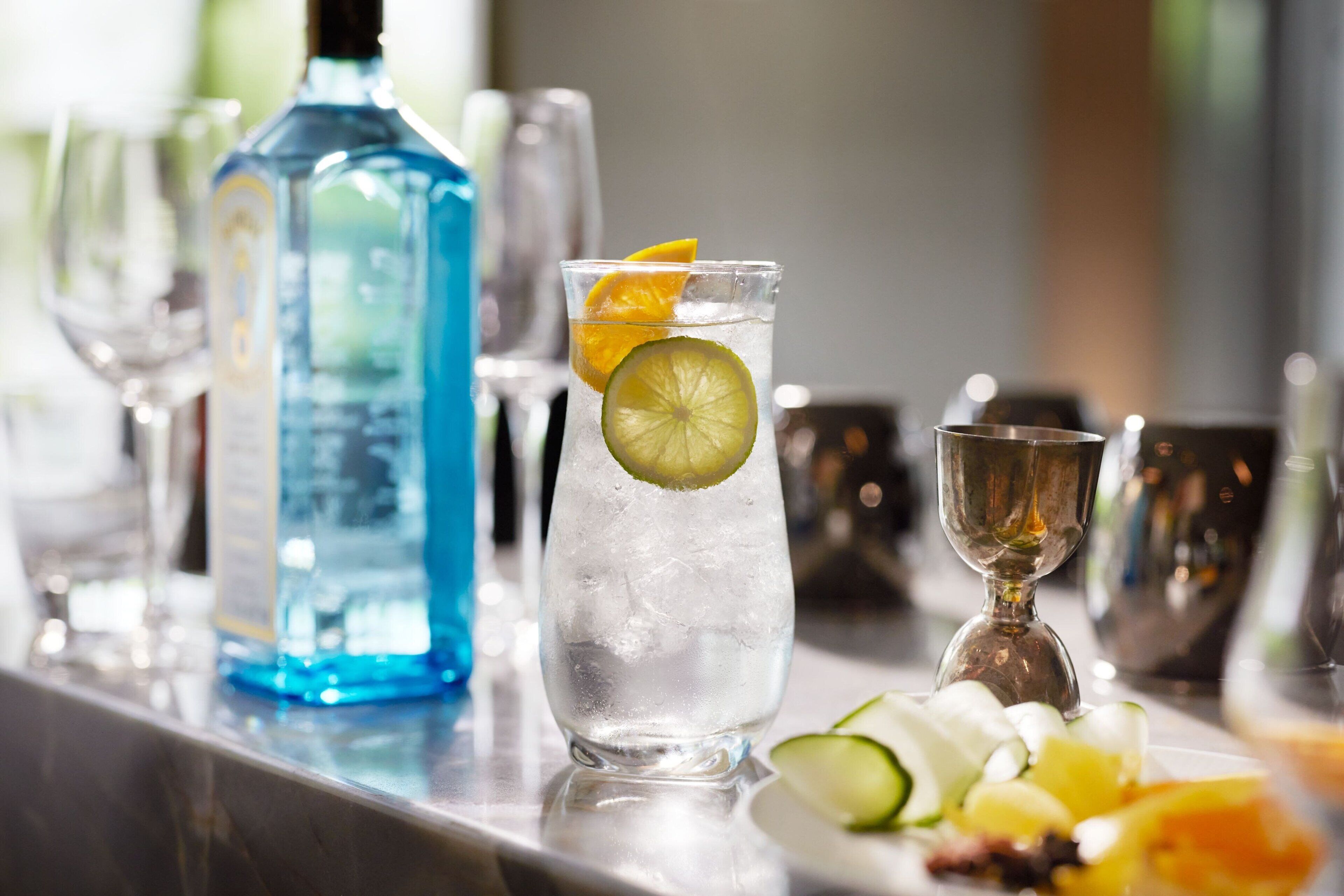 Um balcão de bar com um copo de bebida gelada adornado com limão e uma fatia de laranja, uma garrafa de licor azul e uma variedade de frutas no balcão.