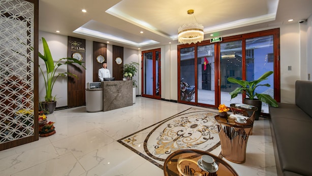 Uma área de recepção de hotel com recepcionista, balcão de atendimento, lustre, placa de recepção, relógio, planta em vaso, sofá e tapete estampado.