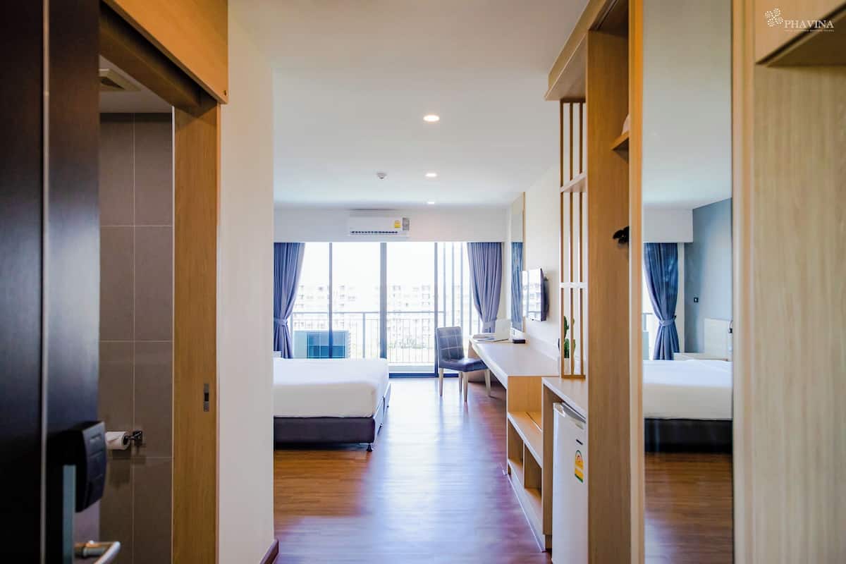 Quarto de hotel com cama, escrivaninha, cadeira e vista da cidade através de grandes janelas.