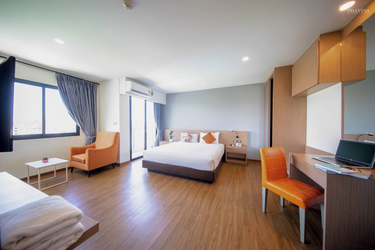 Quarto de hotel moderno com uma cama grande, duas cadeiras, uma escrivaninha com um laptop, uma mesinha de centro com uma planta e uma janela com cortinas.