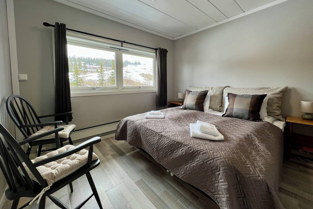 Quarto com cama, uma cadeira, janela com cortinas, mesa de cabeceira e abajur.