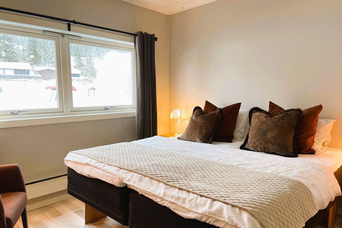 Quarto com cama grande, janela com cortinas, luminária de cabeceira e uma cadeira.