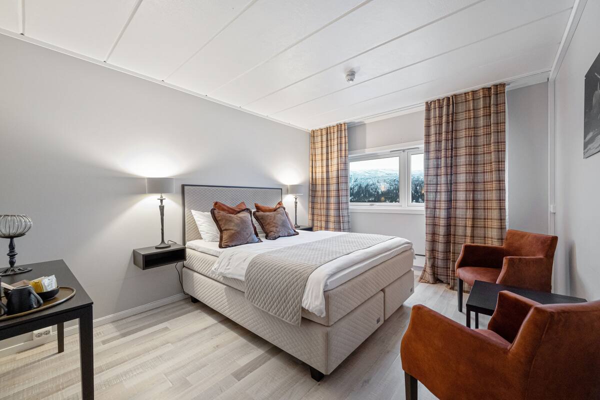Quarto com cama, luminárias de cabeceira, mesa de cabeceira, escrivaninha e cadeira.