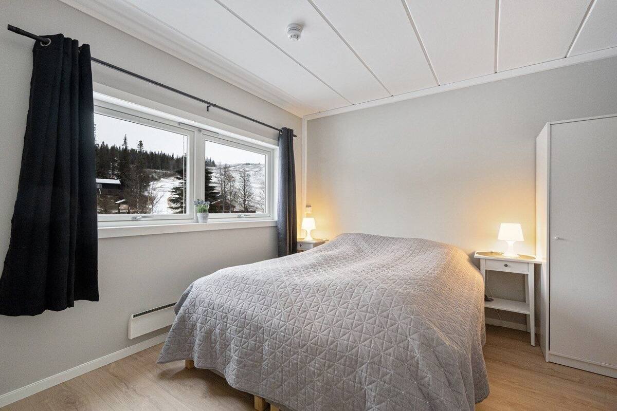 Um quarto com cama, janela com cortinas, mesa de cabeceira com abajur e guarda-roupa.