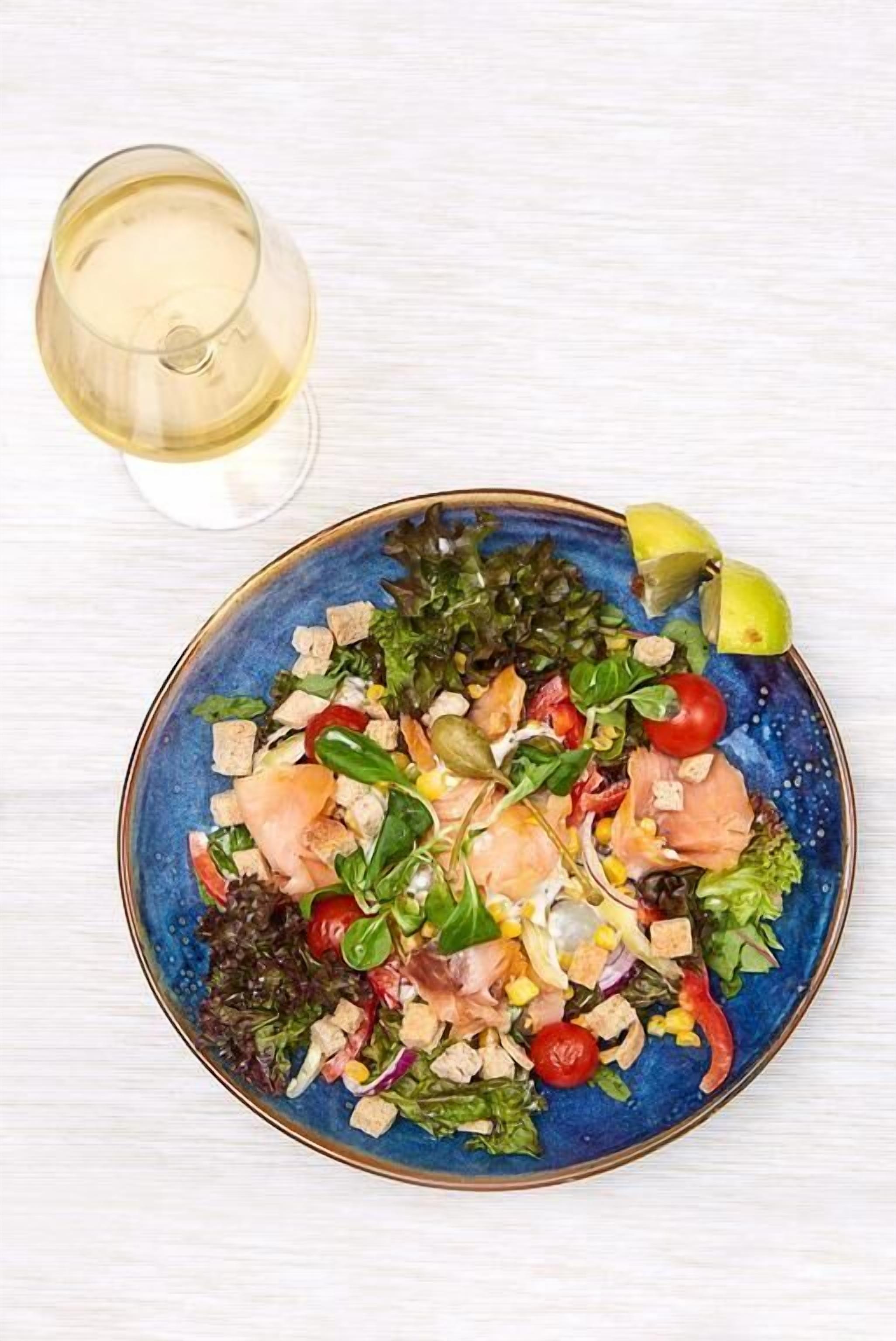 Uma salada com diversos vegetais e um copo de vinho branco sobre uma mesa de madeira.