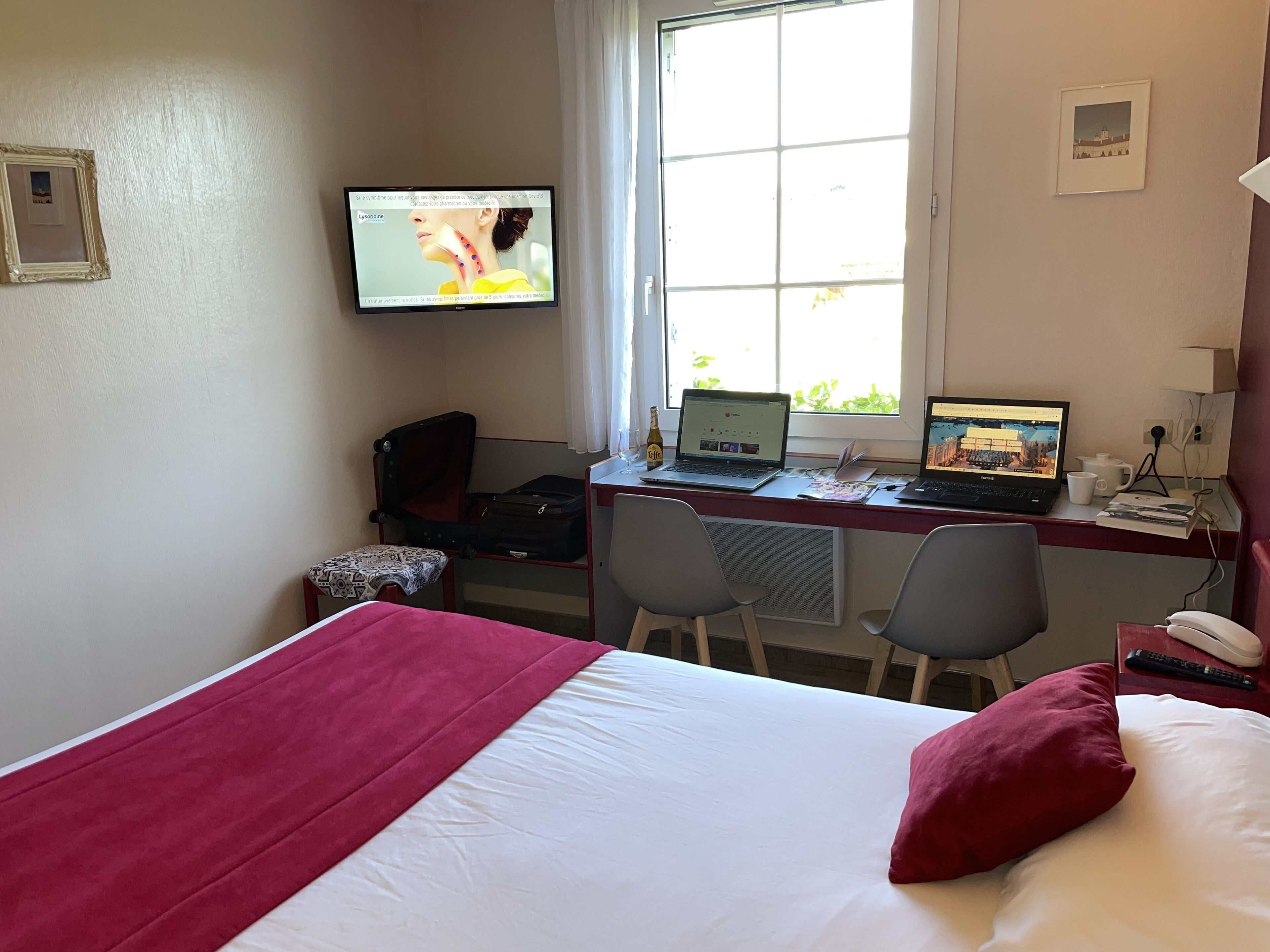 Quarto de hotel com uma cama, uma escrivaninha com dois computadores portáteis, uma cadeira, uma mesinha e uma televisão na parede.