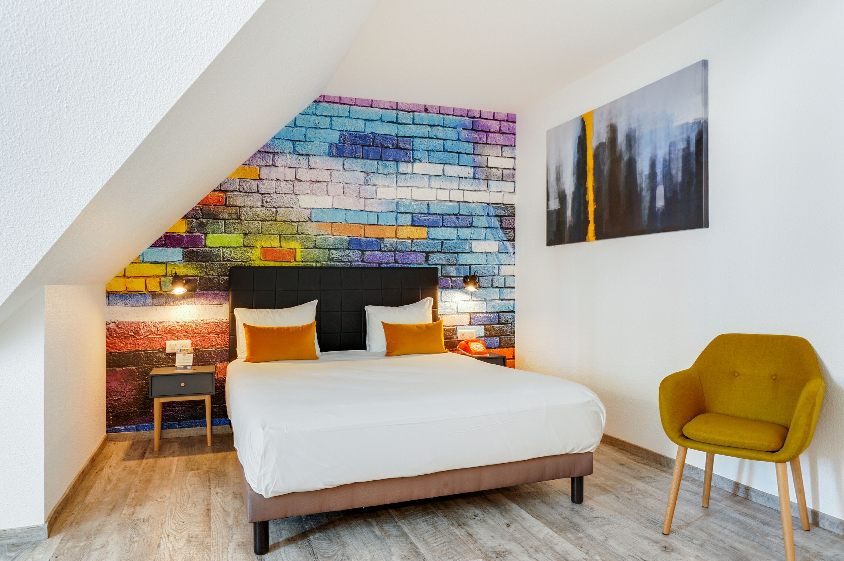 Quarto com teto inclinado, uma cama com roupa de cama branca, uma poltrona amarela e uma parede com padrão de tijolos coloridos.