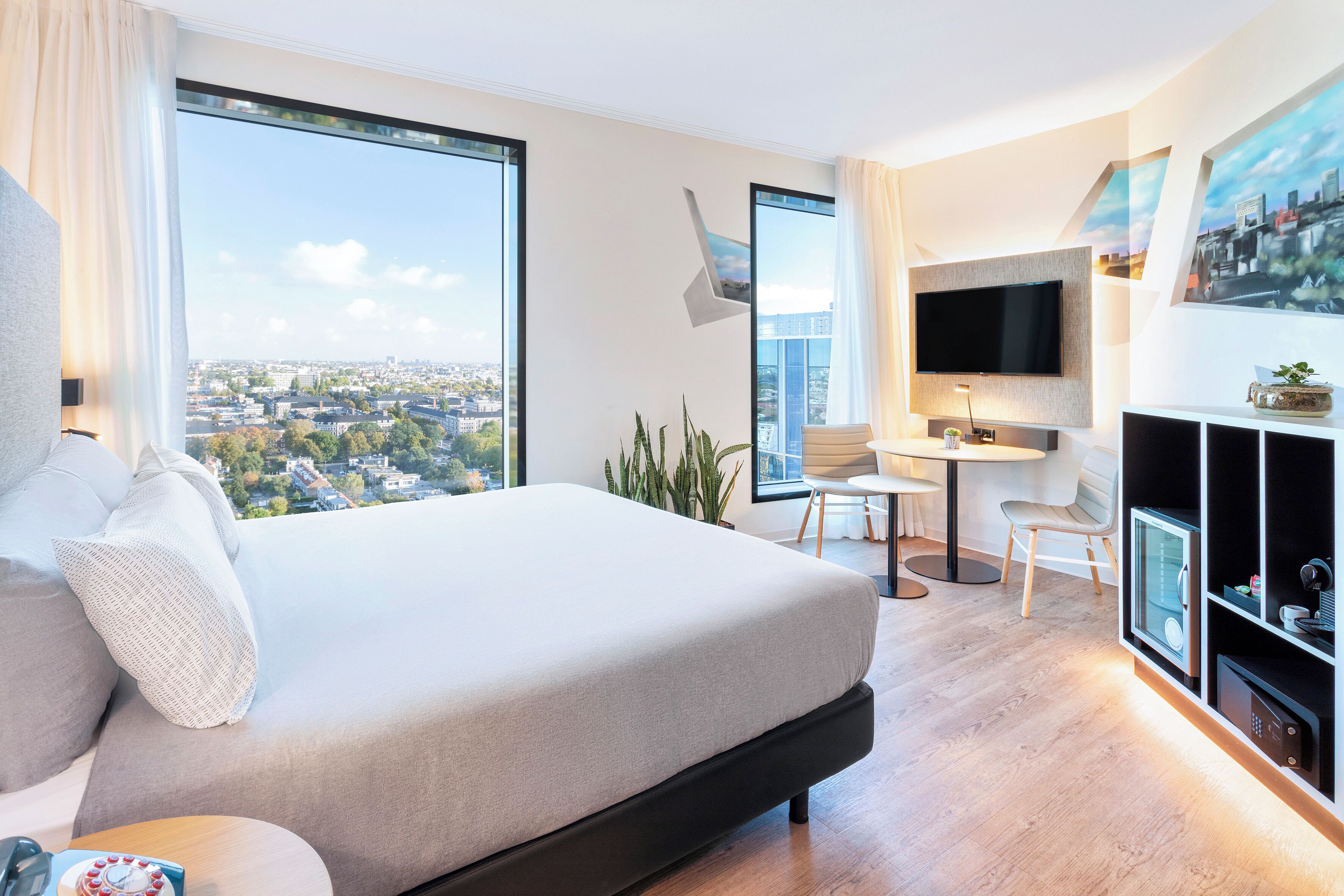 Quarto de hotel moderno com uma cama grande, uma escrivaninha, televisão e vista para a cidade.