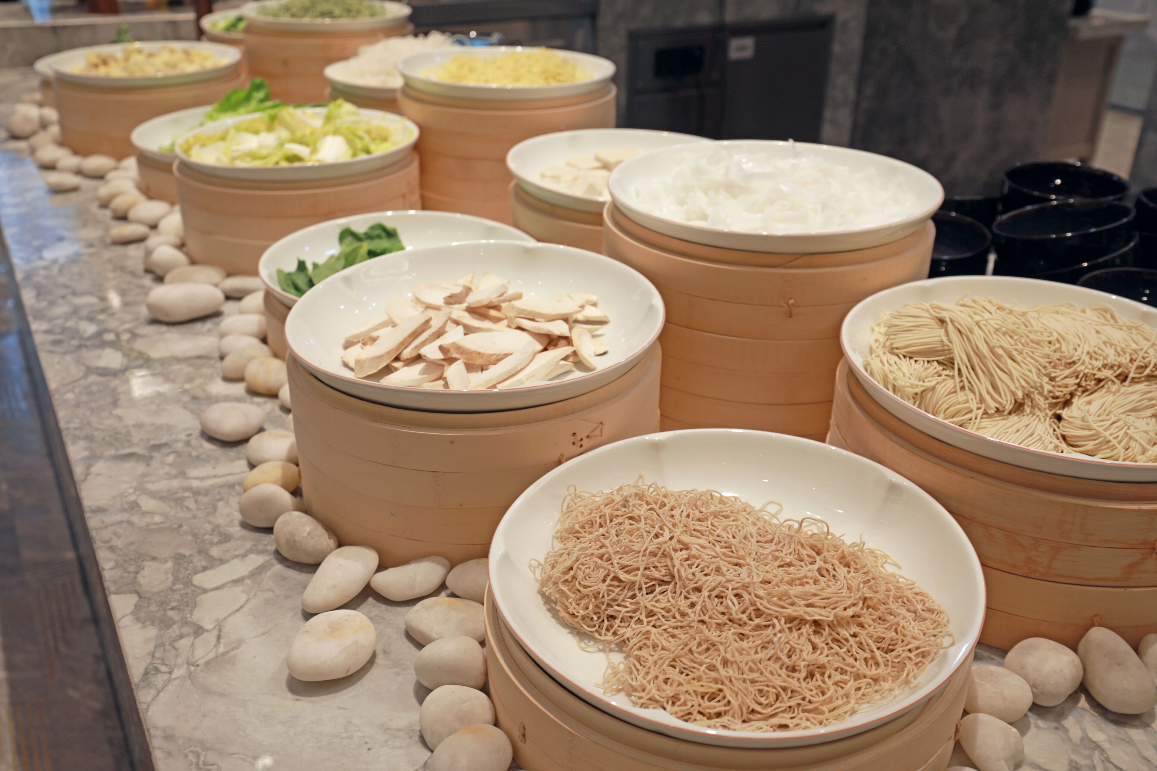 Buffet com diversos alimentos em grandes tigelas e recipientes, dispostos sobre um balcão de mármore com pedrinhas.