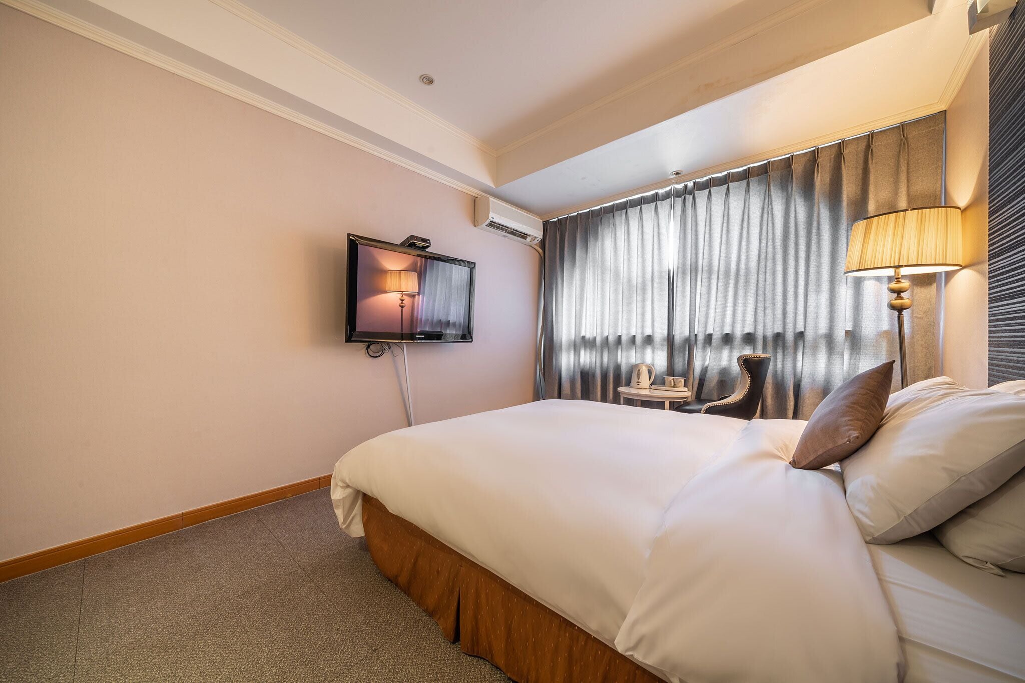 Quarto de hotel com uma cama grande, televisão na parede, uma mesinha com abajur e uma janela com cortinas.