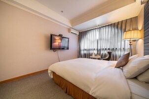Quarto de hotel com uma cama grande, televisão na parede, uma mesinha com abajur e uma janela com cortinas.