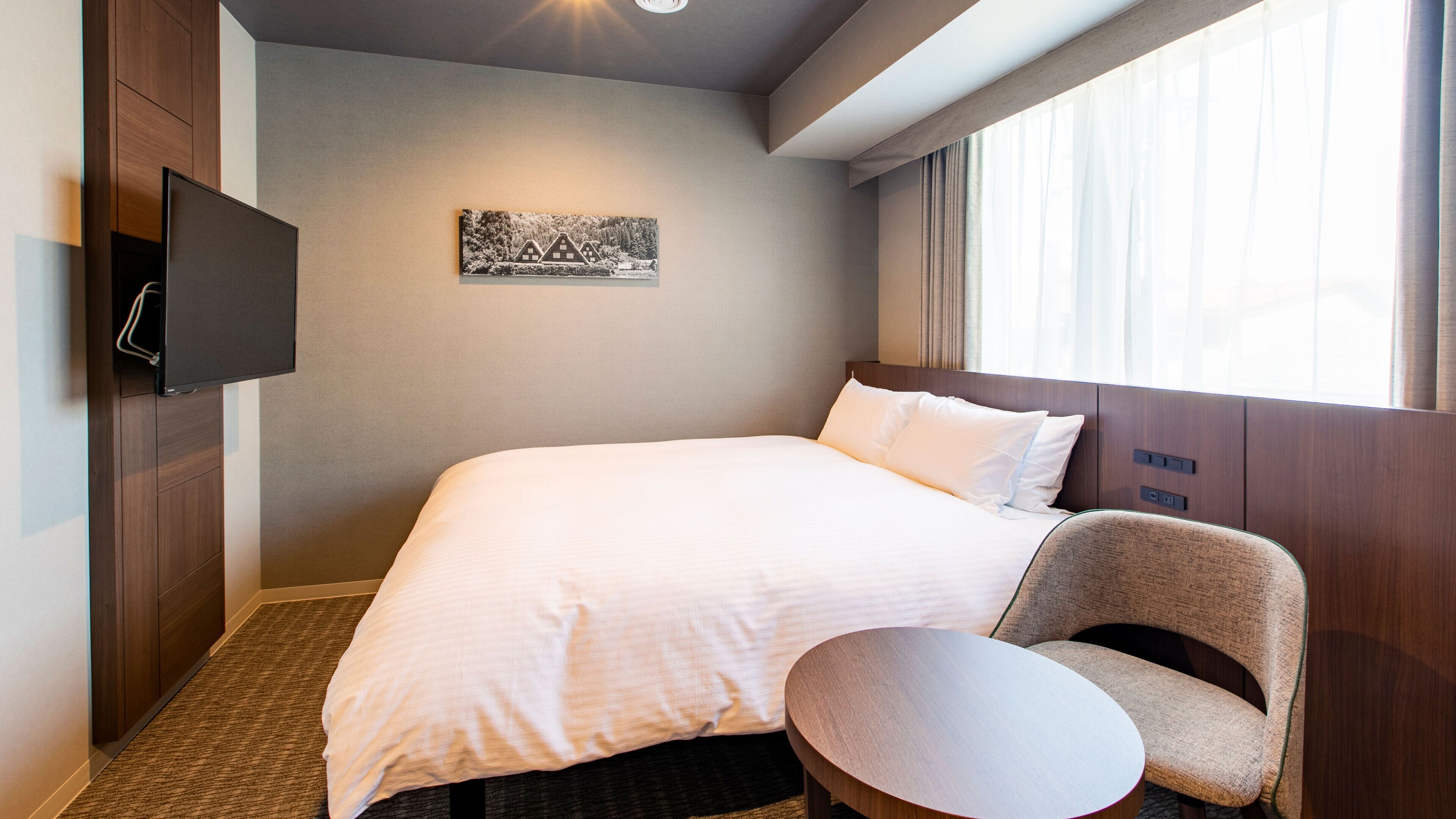 Quarto de hotel com cama, mesa redonda pequena, cadeira, televisão de tela plana na parede e janela com cortinas.
