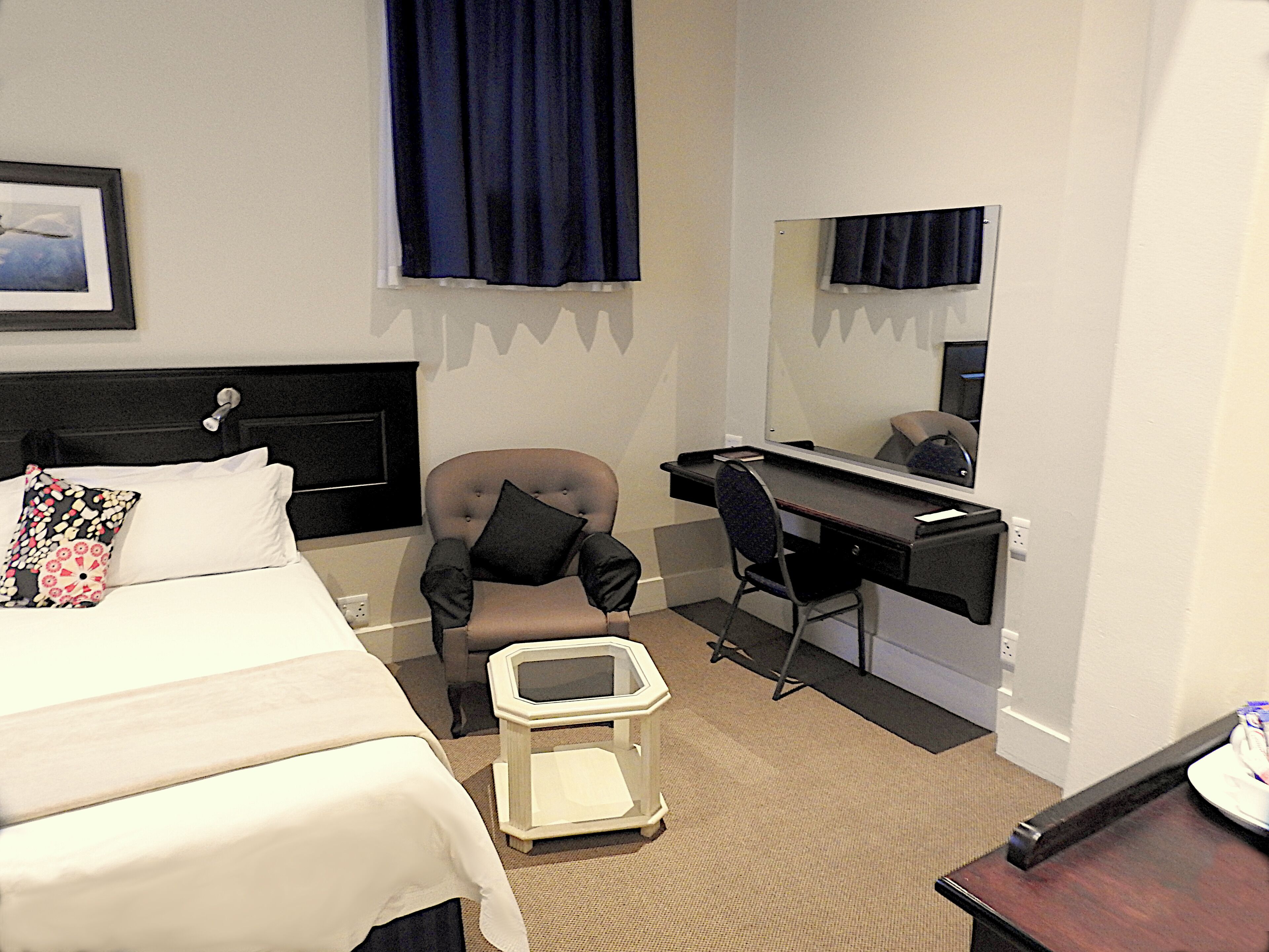 Quarto de hotel com cama, escrivaninha, cadeira, um banquinho pequeno, espelho e janela com cortinas.