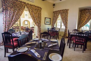 Uma sala de jantar com mesas preparadas para quatro pessoas, cada uma com cadeiras estofadas vermelhas, e cortinas florais.