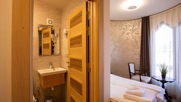 Quarto de hotel com porta de madeira, pia, espelho, cama com toalhas, cadeira, mesa pequena com planta e janela com cortinas.