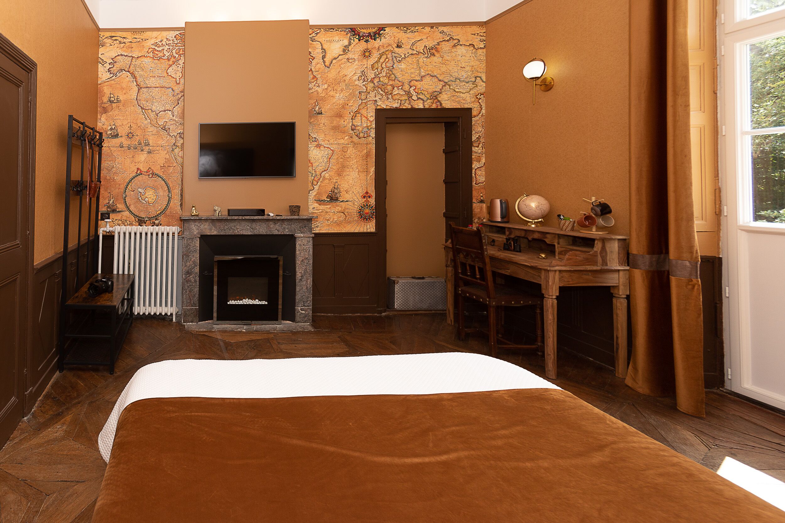 Quarto de hotel com uma cama grande, lareira, escrivaninha com cadeira, televisão na parede e uma janela ampla com cortinas.