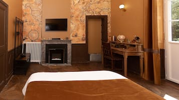 Quarto de hotel com uma cama grande, lareira, escrivaninha com cadeira, televisão na parede e uma janela ampla com cortinas.