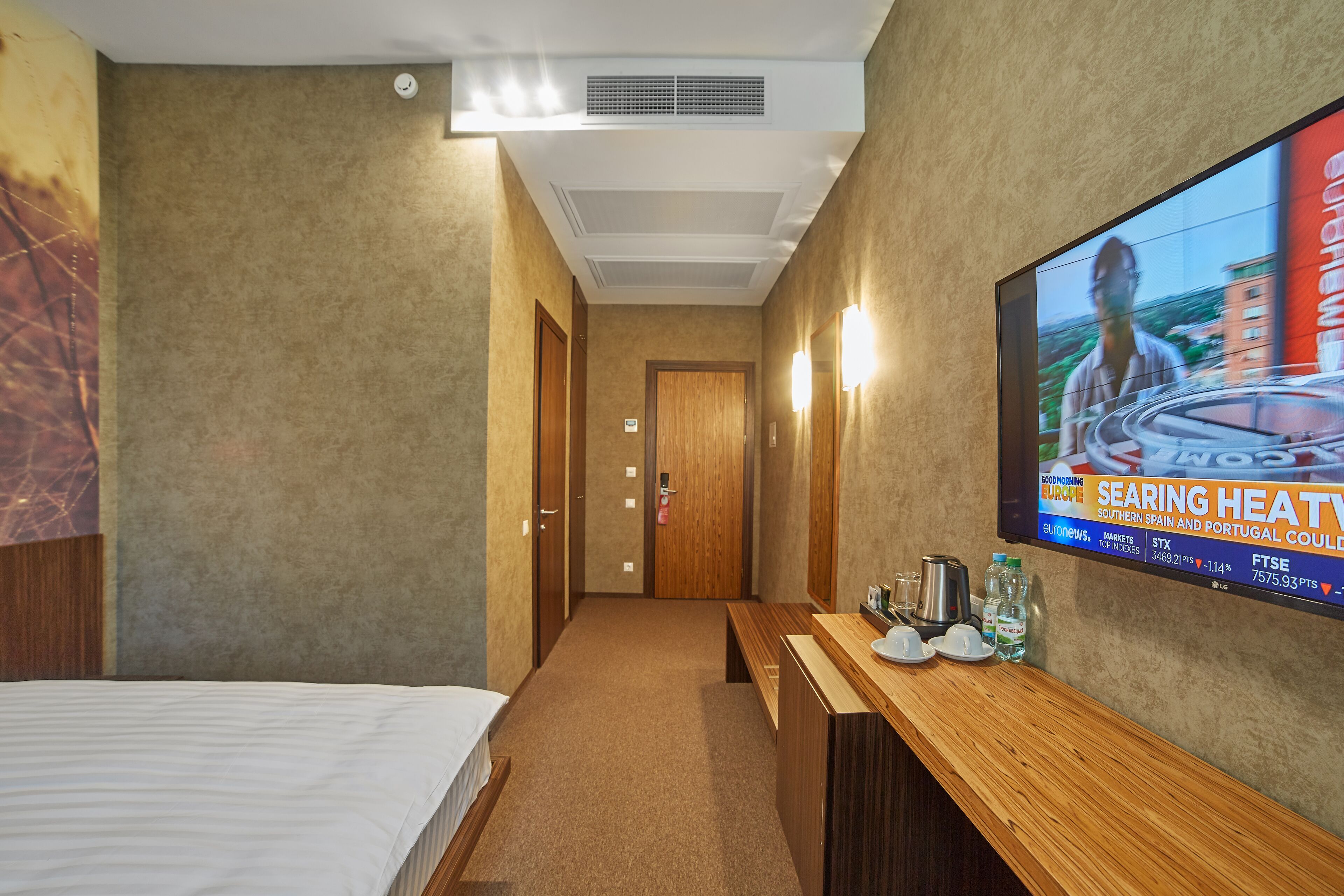 Quarto de hotel com cama, um banco de madeira, uma televisão exibindo um noticiário e uma TV na parede.