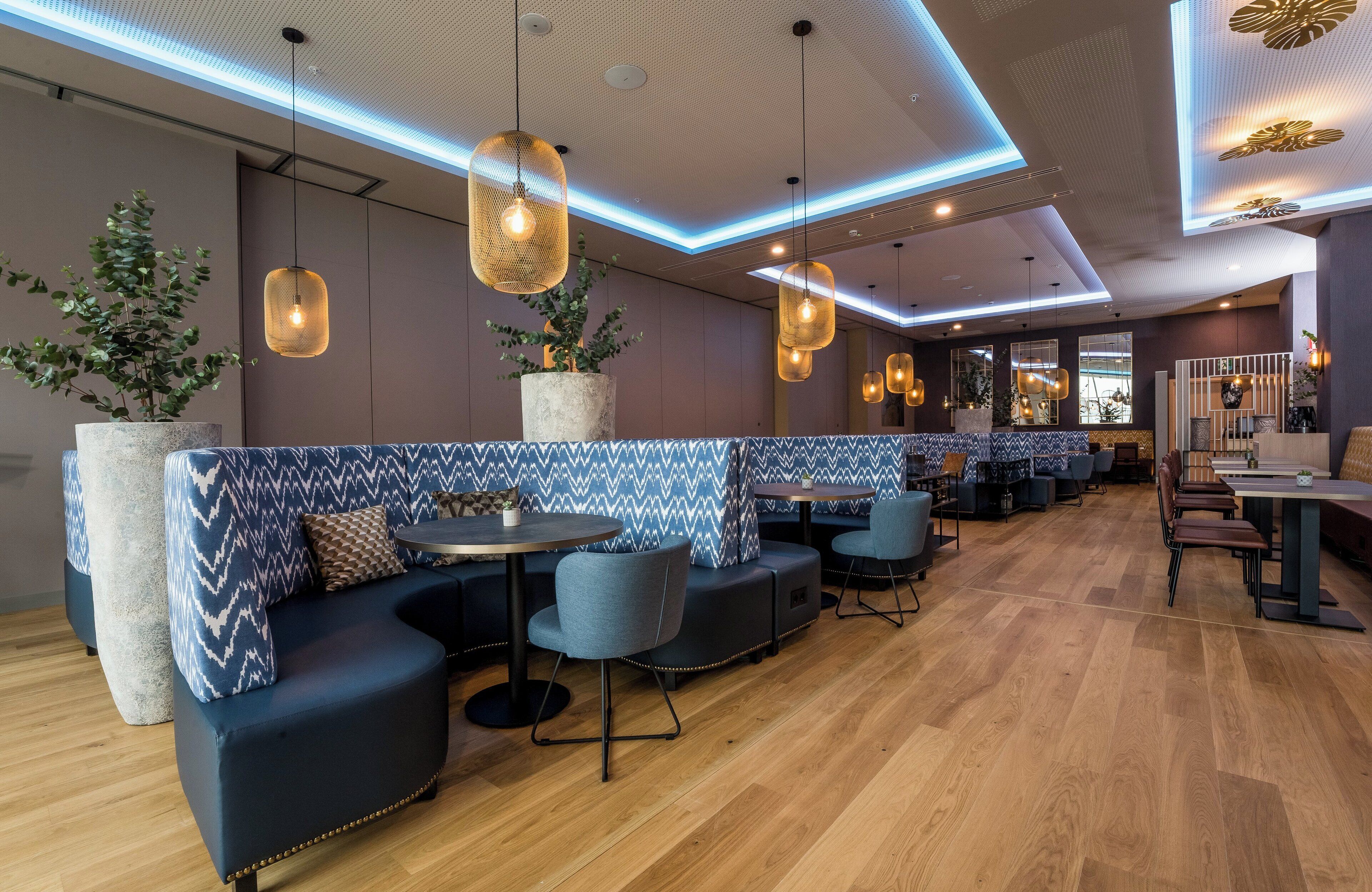 Um restaurante moderno com piso de madeira, assentos estampados em azul e branco, luminárias pendentes e plantas em vasos.
