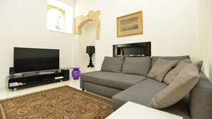 Uma sala de estar com um sofá cinzento, uma televisão de tela plana, um tapete estampado, um pequeno vaso roxo e um quadro na parede.