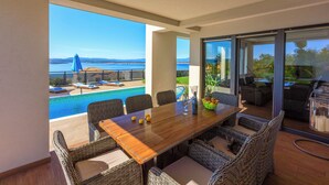 Uma área de refeições com mesa de madeira e cadeiras de vime, com vista para uma piscina e uma paisagem marítima.