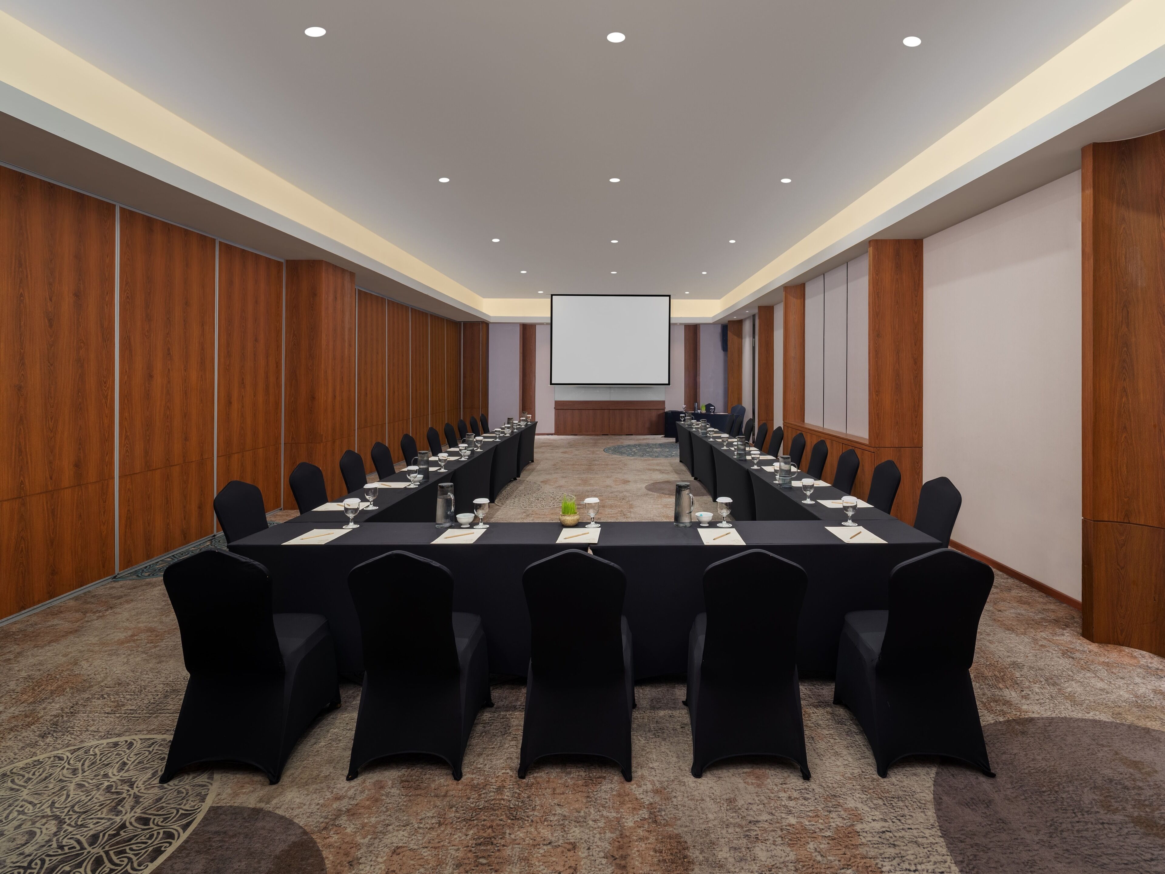 Uma sala de conferências com uma mesa comprida, cadeiras pretas e uma tela de projetor.