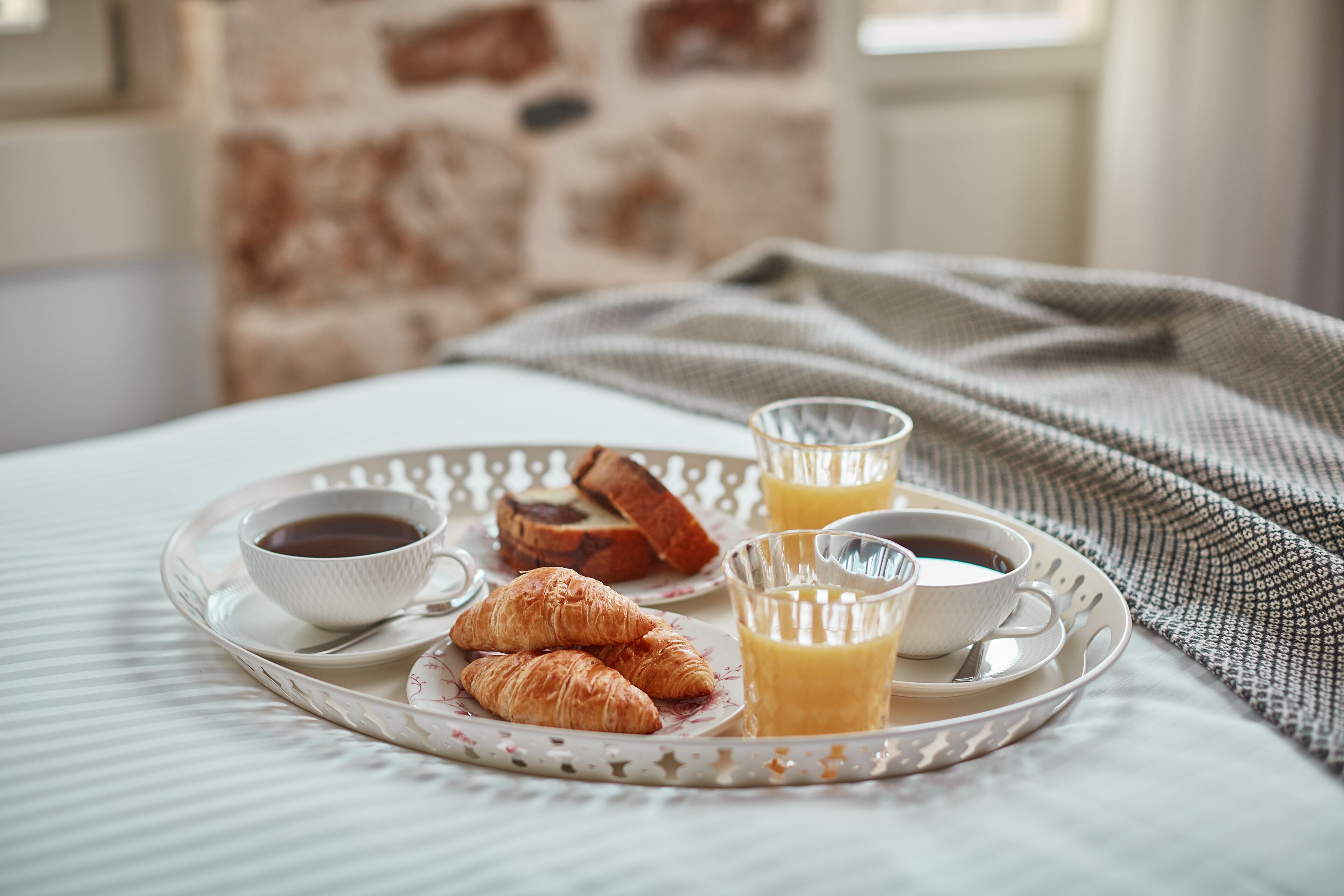 Bandeja de café da manhã com croissants, café e suco de laranja sobre uma cama com colcha listrada.