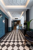 Um hall de hotel com piso xadrez, escada, balcão de recepção e uma grande planta.