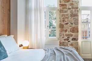 Quarto com cama, luminária de cabeceira, janela com vista para o verde e parede de pedra.