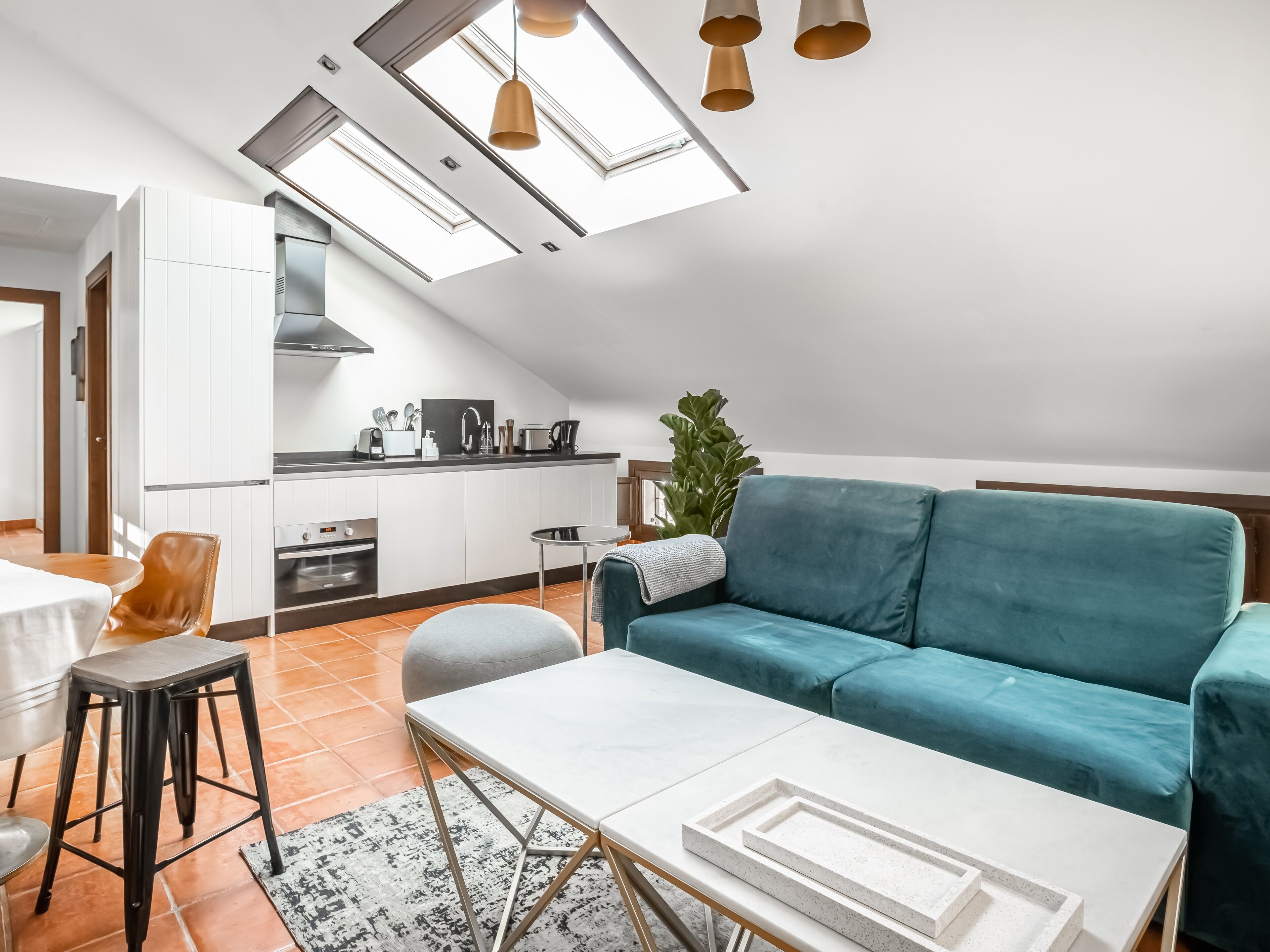 Uma cozinha moderna com claraboias, uma sala de jantar com mesa e cadeiras, e um sofá verde-água numa sala de estar.