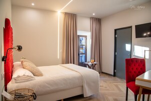Quarto de hotel com cama, uma cadeira vermelha, mesa com copo de suco de laranja e televisão na parede.