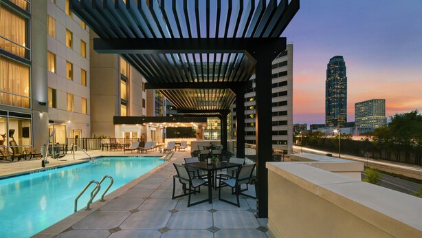 Uma área de piscina no terraço, com um espaço de estar coberto, piscina com guarda-corpo de metal e vista para a cidade ao fundo.