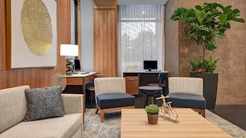 Quarto de hotel moderno com uma janela ampla, uma escrivaninha de madeira, duas poltronas, uma mesinha de centro e uma planta grande.