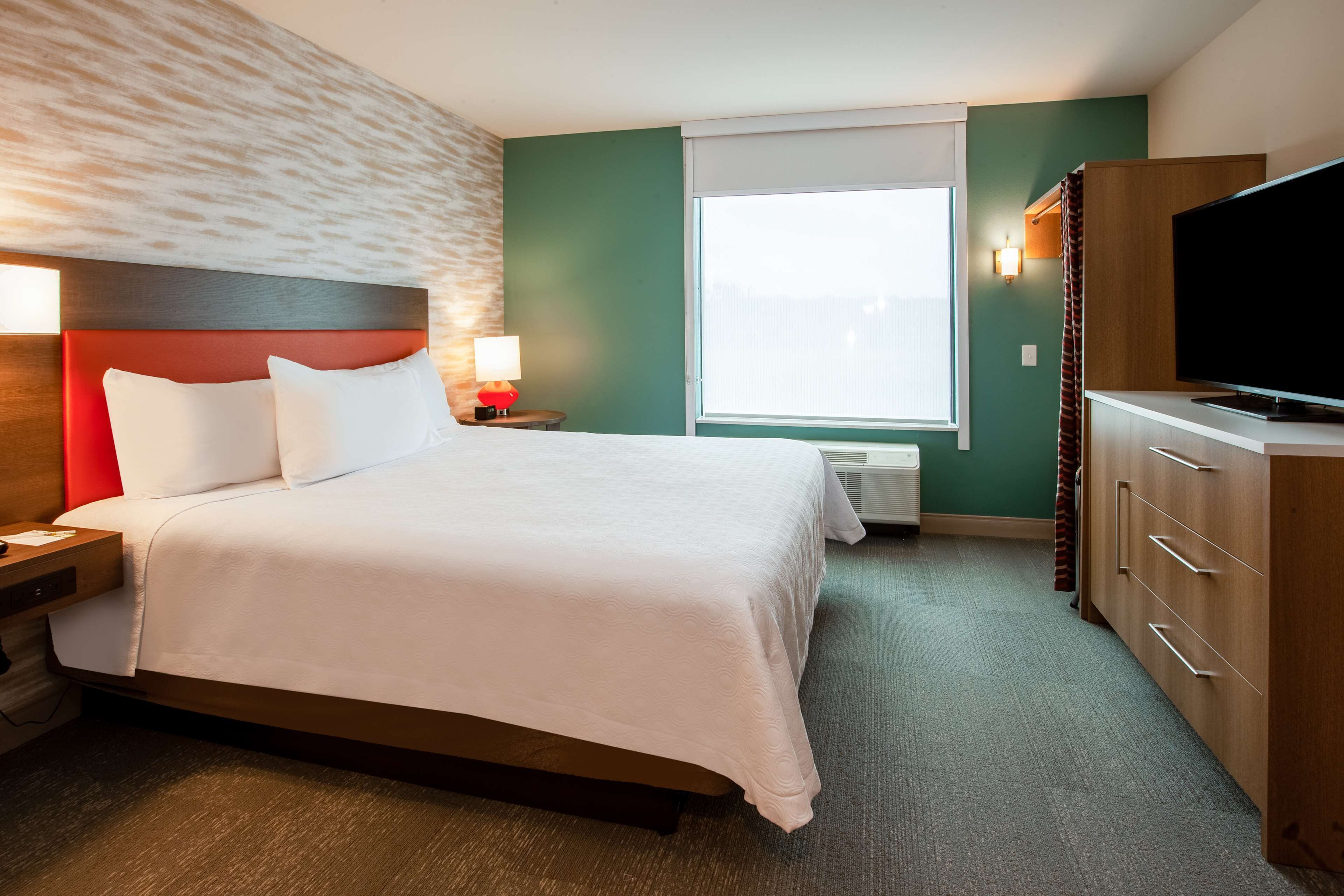 Quarto de hotel com cama, televisão, mesa de cabeceira e janela com persianas.