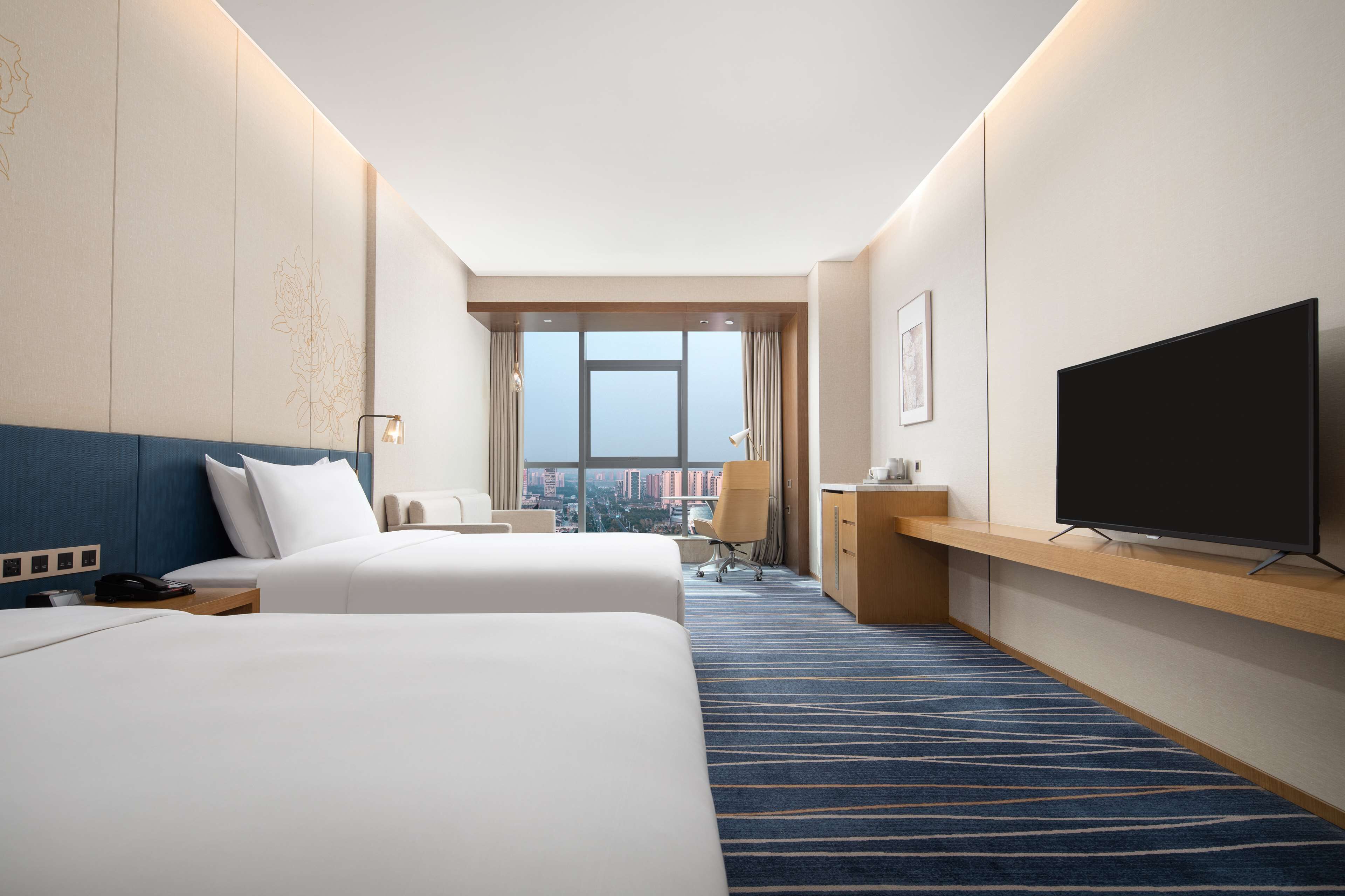 Quarto de hotel com duas camas, televisão, secretária e vista para a cidade.