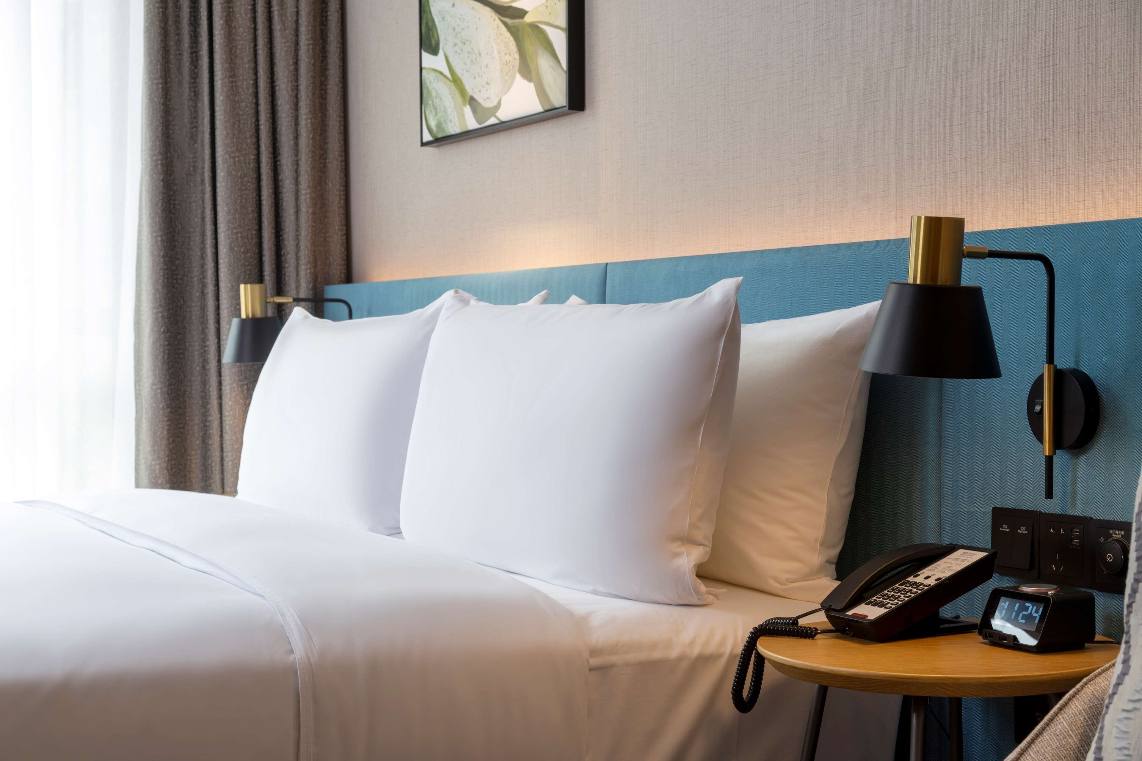 Quarto de hotel com cama, mesa de cabeceira com telefone e relógio digital, luminária na parede e um quadro.