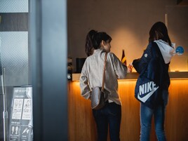 Duas pessoas no balcão de recepção de um hotel, com um balcão de madeira e uma tela digital exibindo informações.